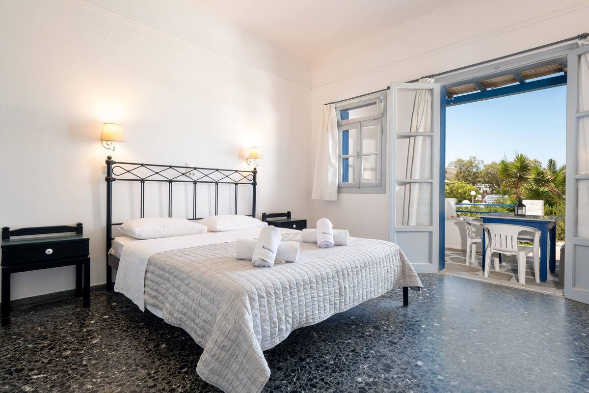 Quarto de hotel com cama, duas mesas de cabeceira, escrivaninha com cadeiras e varanda com vista.