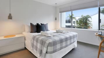 Quarto moderno com cama, mesas de cabeceira, janela com vista para palmeiras e ventilador de teto.