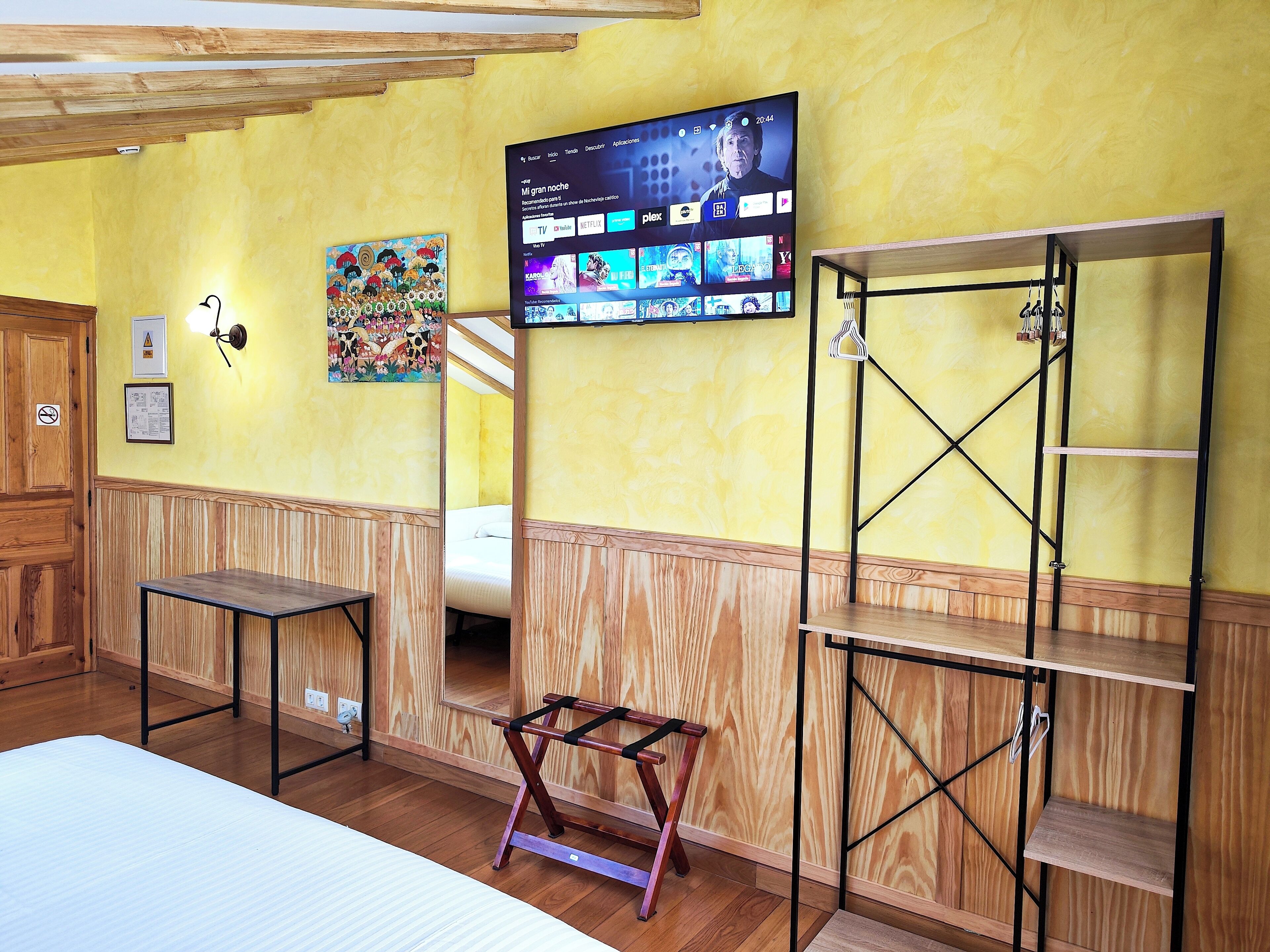 Quarto com painéis de madeira, TV na parede exibindo diversos serviços de streaming, uma pequena mesa de madeira e uma estante de metal.