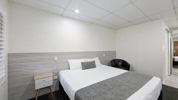 Quarto de hotel com uma cama grande, uma mesinha de cabeceira, uma cadeira e vista para o corredor.