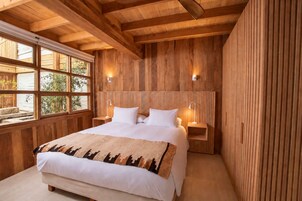 Quarto de madeira com uma cama grande, duas mesas de cabeceira com abajures e uma janela com vista para o verde.