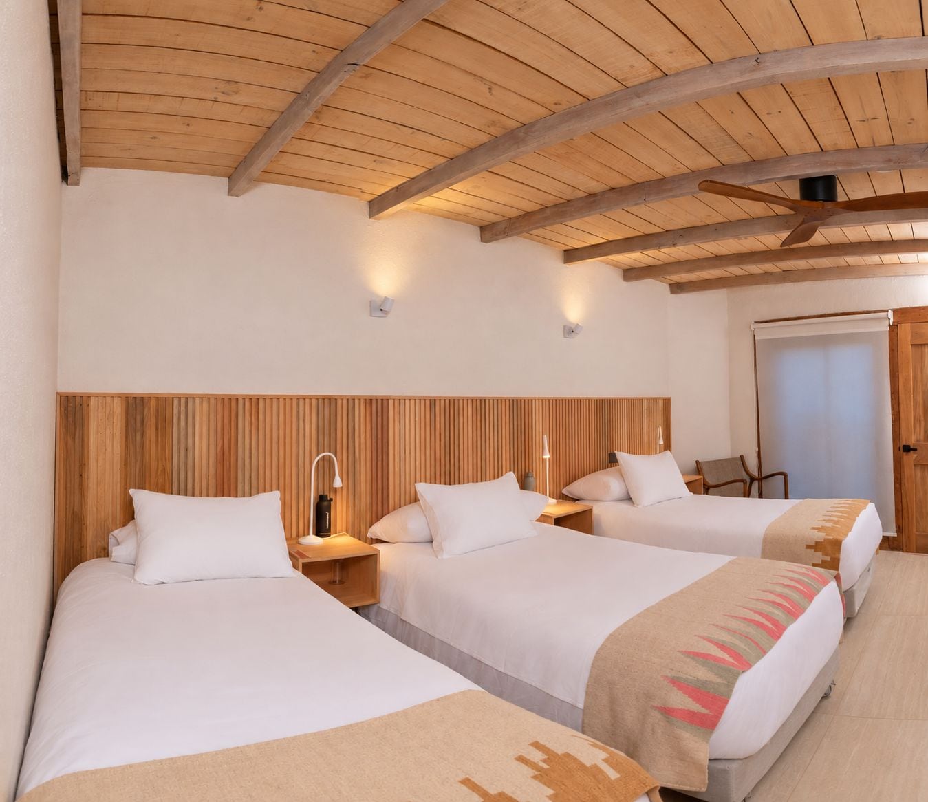 Quarto de hotel com três camas e cabeceiras de madeira, além de teto inclinado.