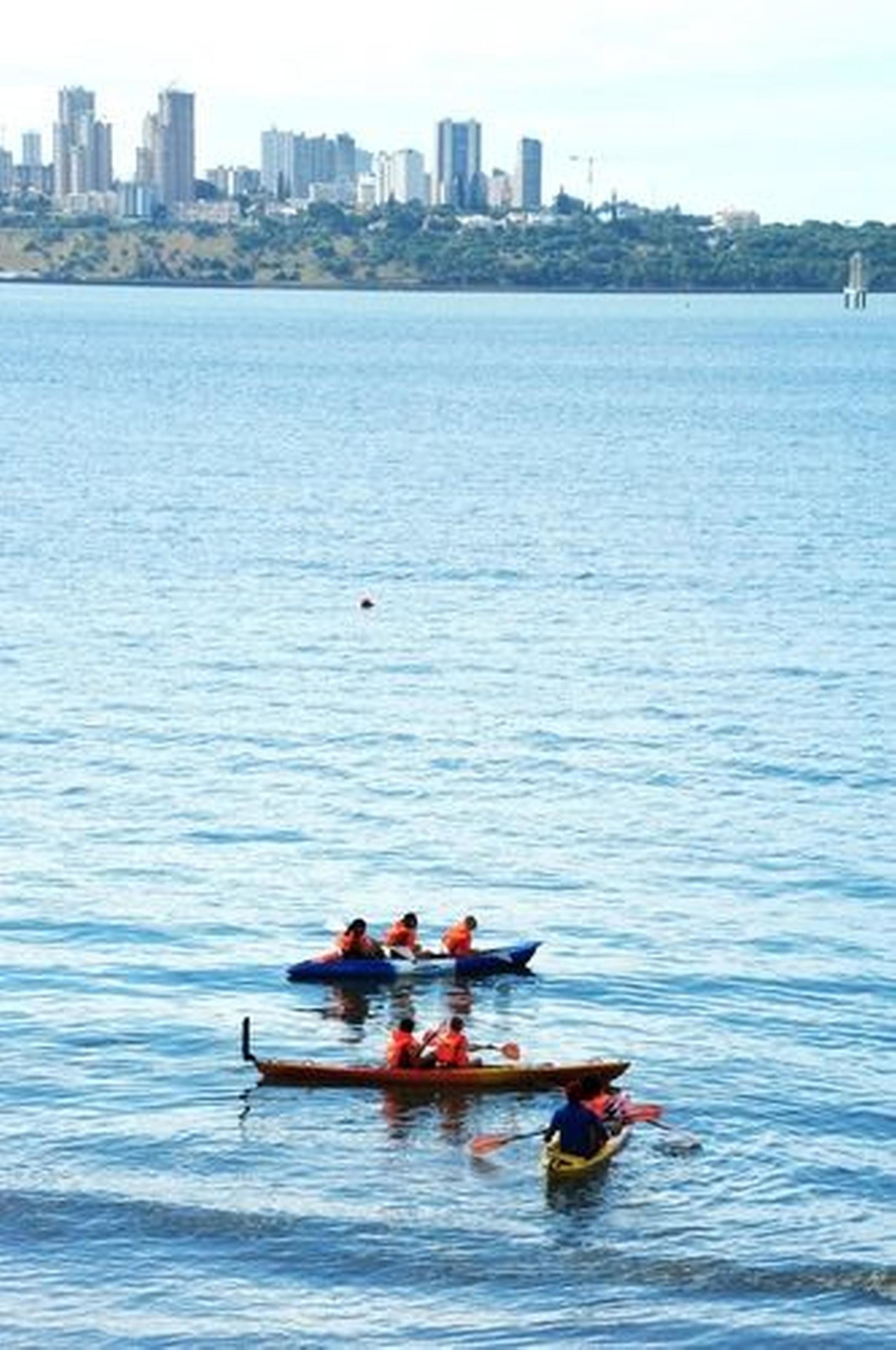 Um grupo de pessoas remando em águas calmas, com o horizonte da cidade ao fundo.
