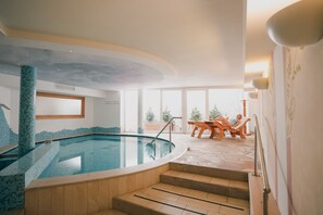 Piscina interior com deck de madeira e degraus dando acesso à piscina, cercada por amplas janelas e vista para árvores.