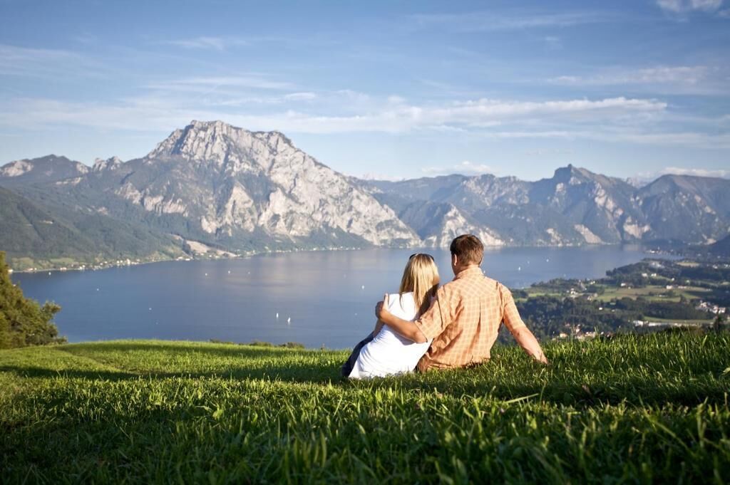 Um casal sentado numa colina gramada com vista para um lago e montanhas.