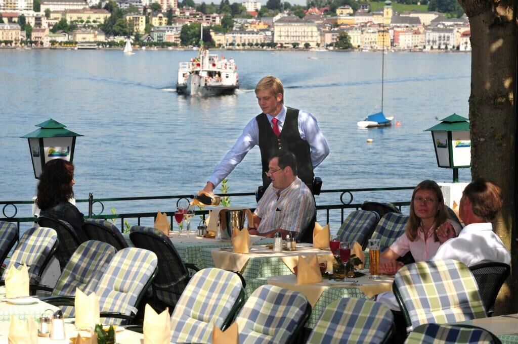 Um restaurante à beira-mar com mesas ao ar livre, um garçom atendendo clientes e um barco ao fundo.
