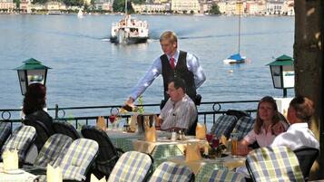 Um restaurante à beira-mar com mesas ao ar livre, um garçom atendendo clientes e um barco ao fundo.