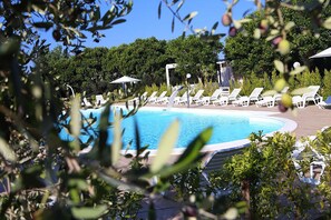 Uma piscina cercada por cadeiras de descanso brancas e guarda-sóis, com árvores ao fundo.