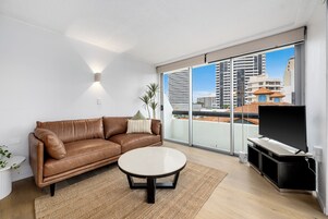 Uma sala de estar moderna com um sofá de couro marrom, uma mesa de centro redonda e uma janela grande com vista para edifícios altos.
