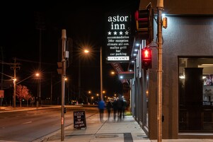 Vista noturna da rua com um letreiro do 'Astor Inn' anunciando comodidades como hospedagem, recepção 24 horas, controle de temperatura, um lounge e um bar.