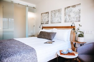 Quarto de hotel com uma cama, uma mesinha de cabeceira com uma chávena de café e uma cadeira.