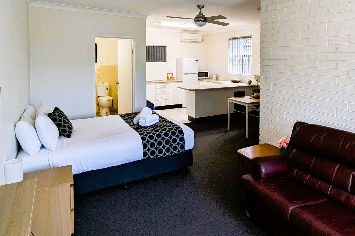 Quarto de hotel com cama, sofá de couro vermelho, escrivaninha com computador e cozinha compacta.