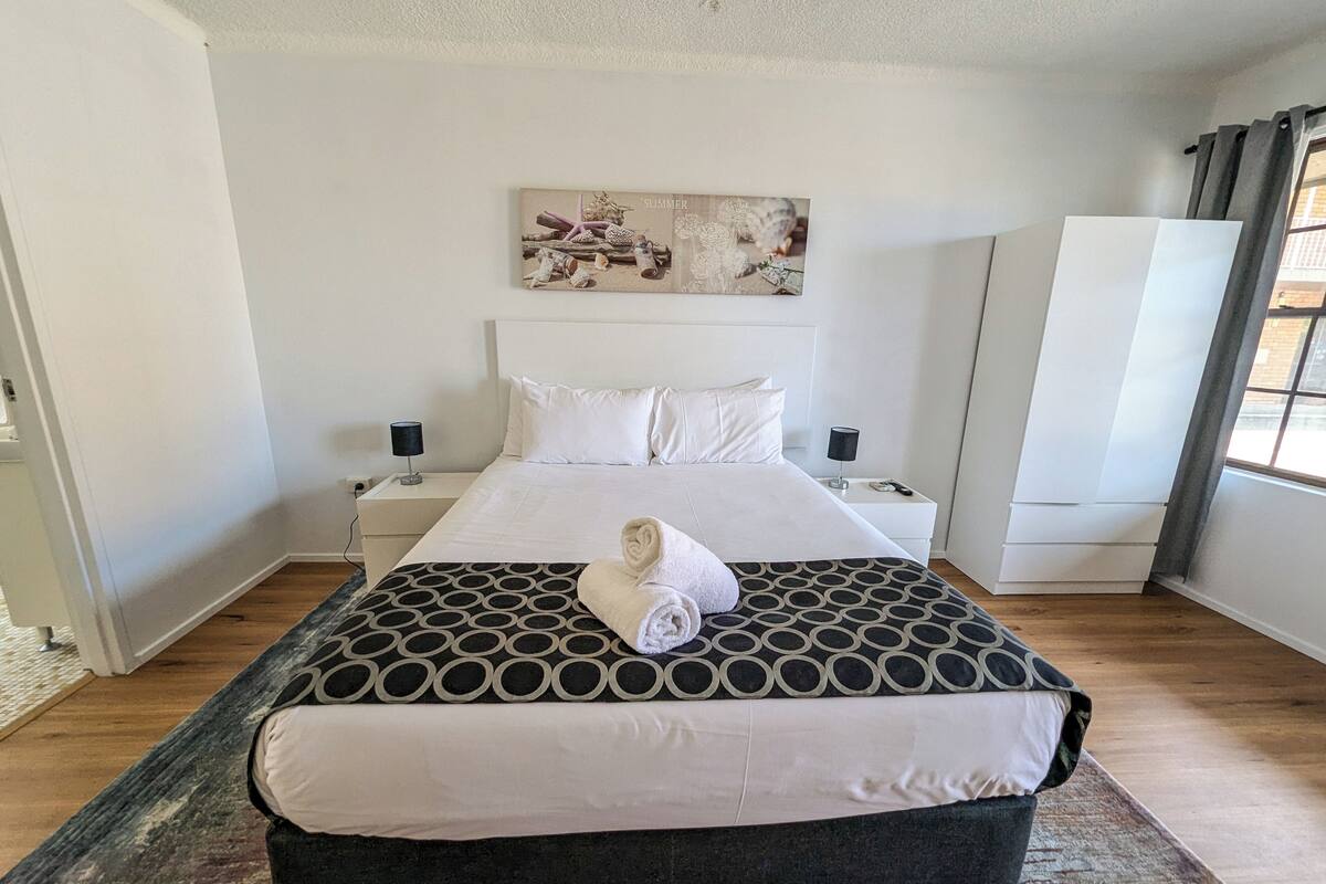 Quarto bem arrumado com uma cama grande, duas mesas de cabeceira, uma obra de arte na parede e um guarda-roupa.