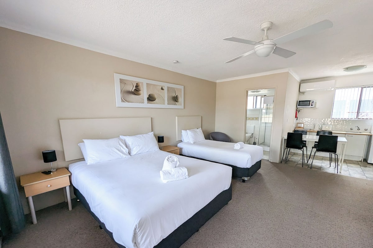 Quarto de hotel com duas camas, um ventilador de teto, uma pequena escrivaninha com cadeira e uma cozinha compacta.