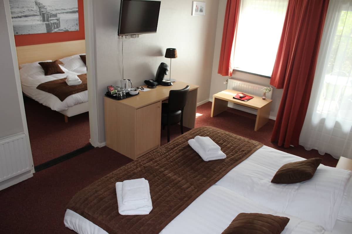Quarto de hotel com duas camas, uma escrivaninha, uma cadeira, uma televisão e uma mesinha de cabeceira com um livro e um abajur.
