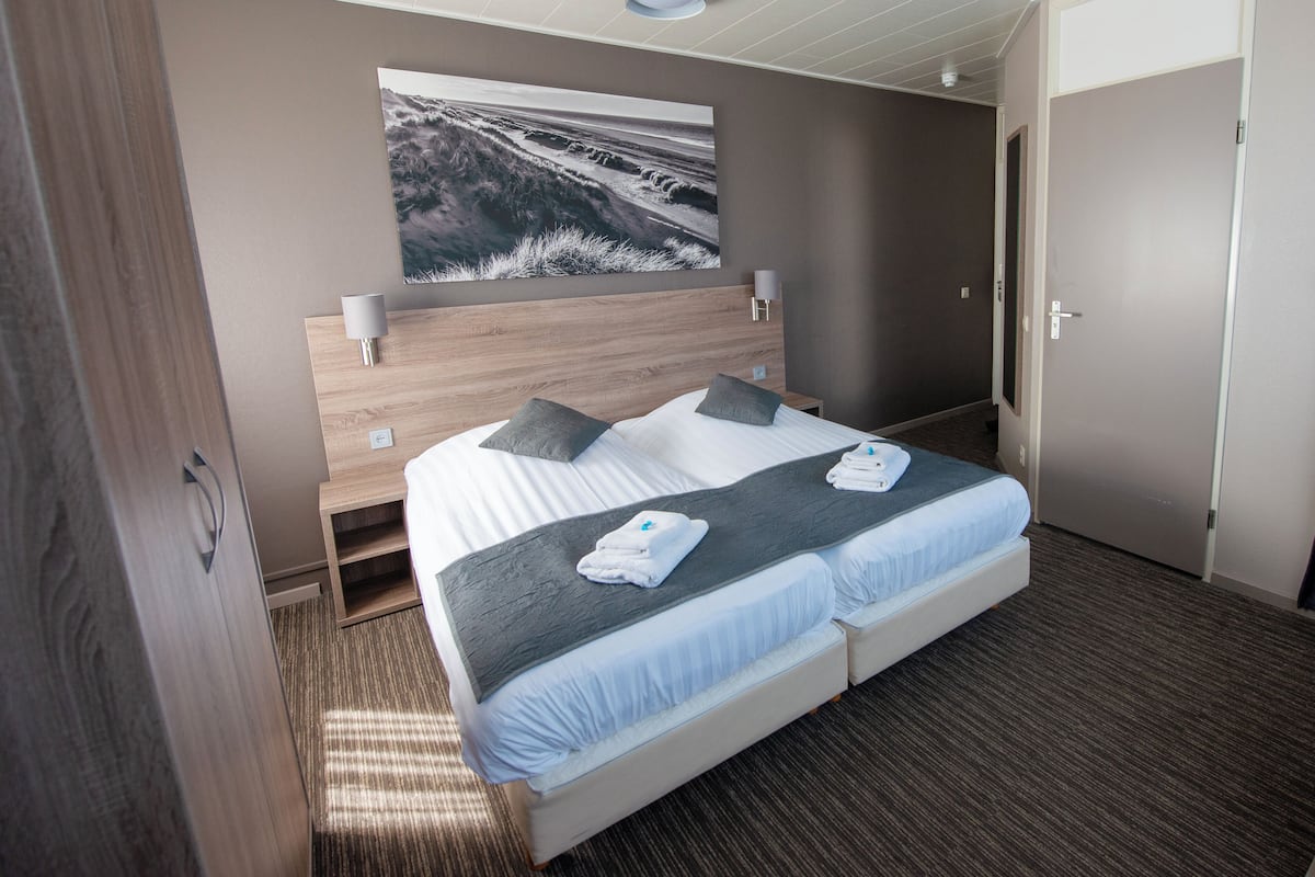 Quarto de hotel com uma cama grande, duas mesas de cabeceira, uma obra de arte na parede e uma porta.