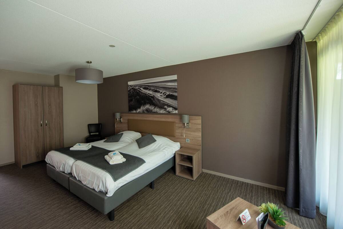 Quarto de hotel moderno com uma cama grande, um guarda-roupa de madeira, uma mesa de cabeceira com abajur, um quadro na parede e uma pequena mesa com uma planta.