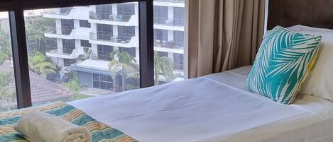 Quarto de hotel com uma cama grande, duas almofadas, um cobertor listrado e vista para um edifício através de uma janela com cortinas.