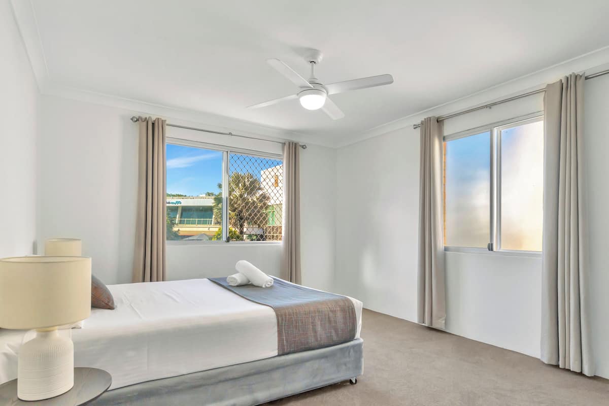 Quarto de hotel com cama, ventilador de teto, janela com cortinas e vista para prédios.