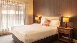 Uma cama bem arrumada com roupa de cama branca e uma almofada decorativa, com duas mesas de cabeceira com abajures, em um quarto com painéis de madeira e cortinas leves.