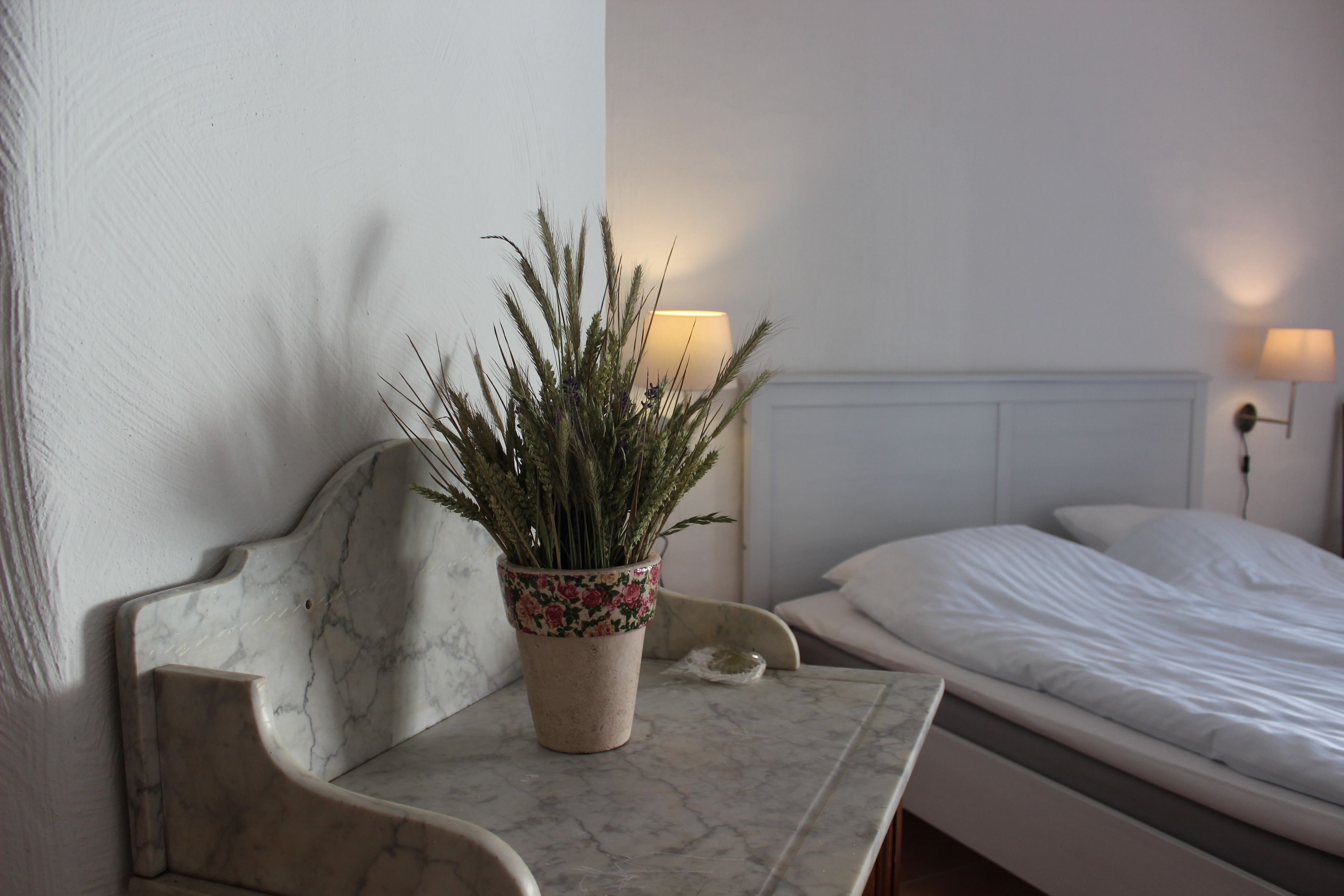 Uma cama com roupa de cama branca, uma mesa de cabeceira de mármore, uma planta em vaso e um abajur fixado na parede.