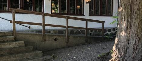 Um edifício com telhado de telhas vermelhas, uma escadaria de madeira que dá para uma área de cascalho, e uma árvore à direita.