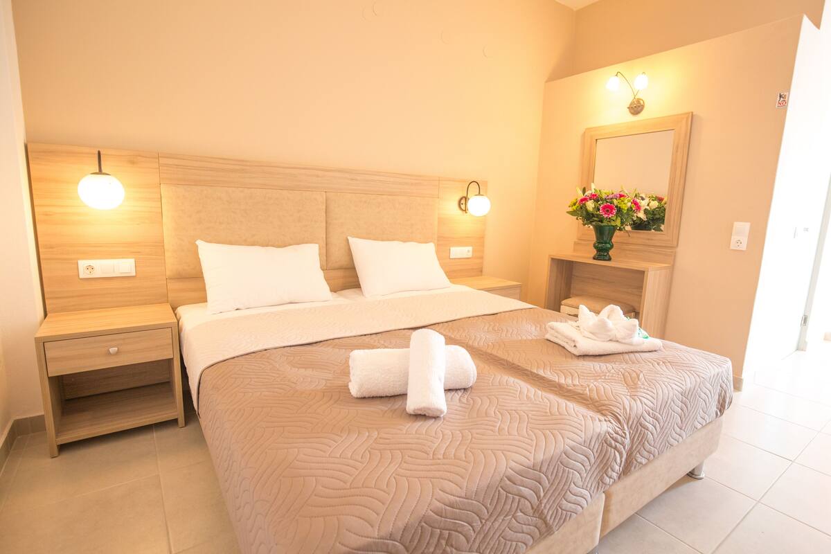 Quarto de hotel com uma cama grande, duas mesas de cabeceira, um criado-mudo, um espelho e um vaso com flores.