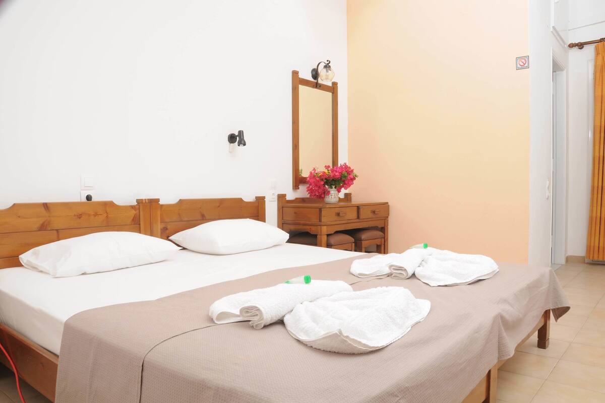 Uma cama de casal com roupa de cama branca, uma cabeceira de madeira, uma mesa de cabeceira com espelho e um vaso com flores.