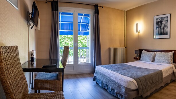 Quarto de hotel com cama, poltrona, mesa, janela com cortinas, televisão na parede e um quadro na parede.