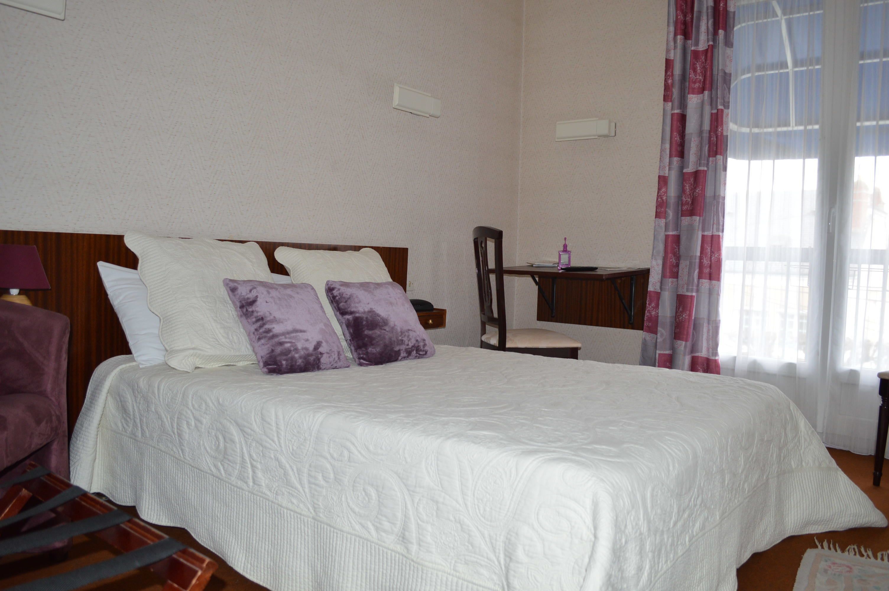 Quarto de hotel com uma cama grande, cabeceira de madeira, mesa de cabeceira, uma cadeira e uma janela com cortinas.