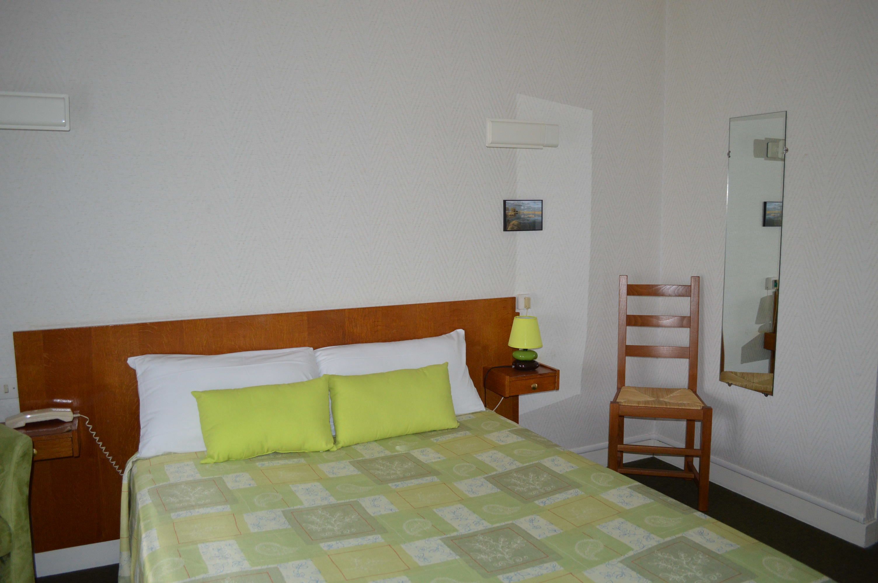 Quarto de hotel com uma cama, mesa de cabeceira, cadeira, luminária, espelho e um pequeno quadro na parede.