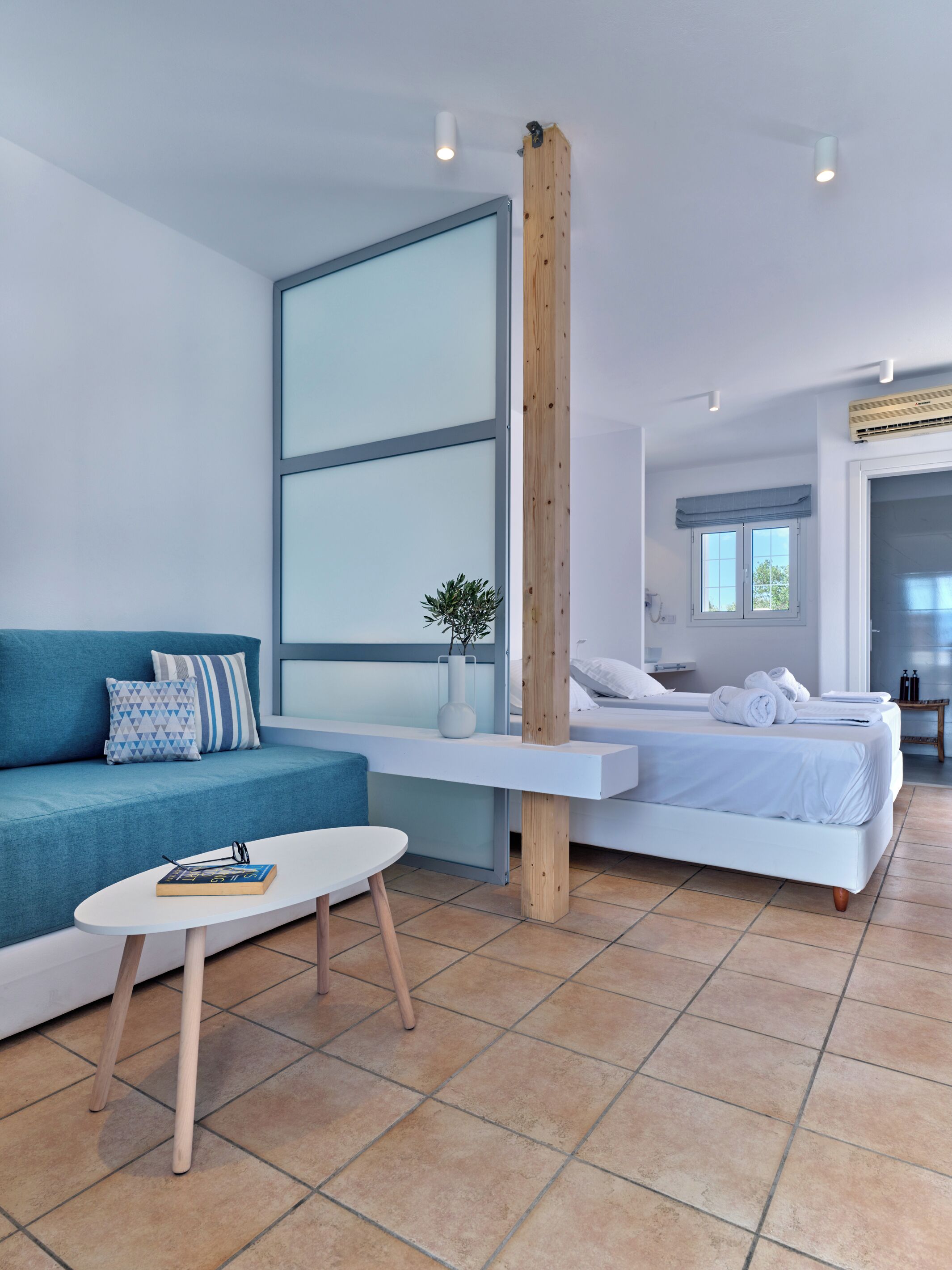 Quarto de hotel moderno com um sofá azul, uma mesa de centro branca, uma parede de destaque em madeira, uma janela ampla e uma cama com roupa de cama branca.