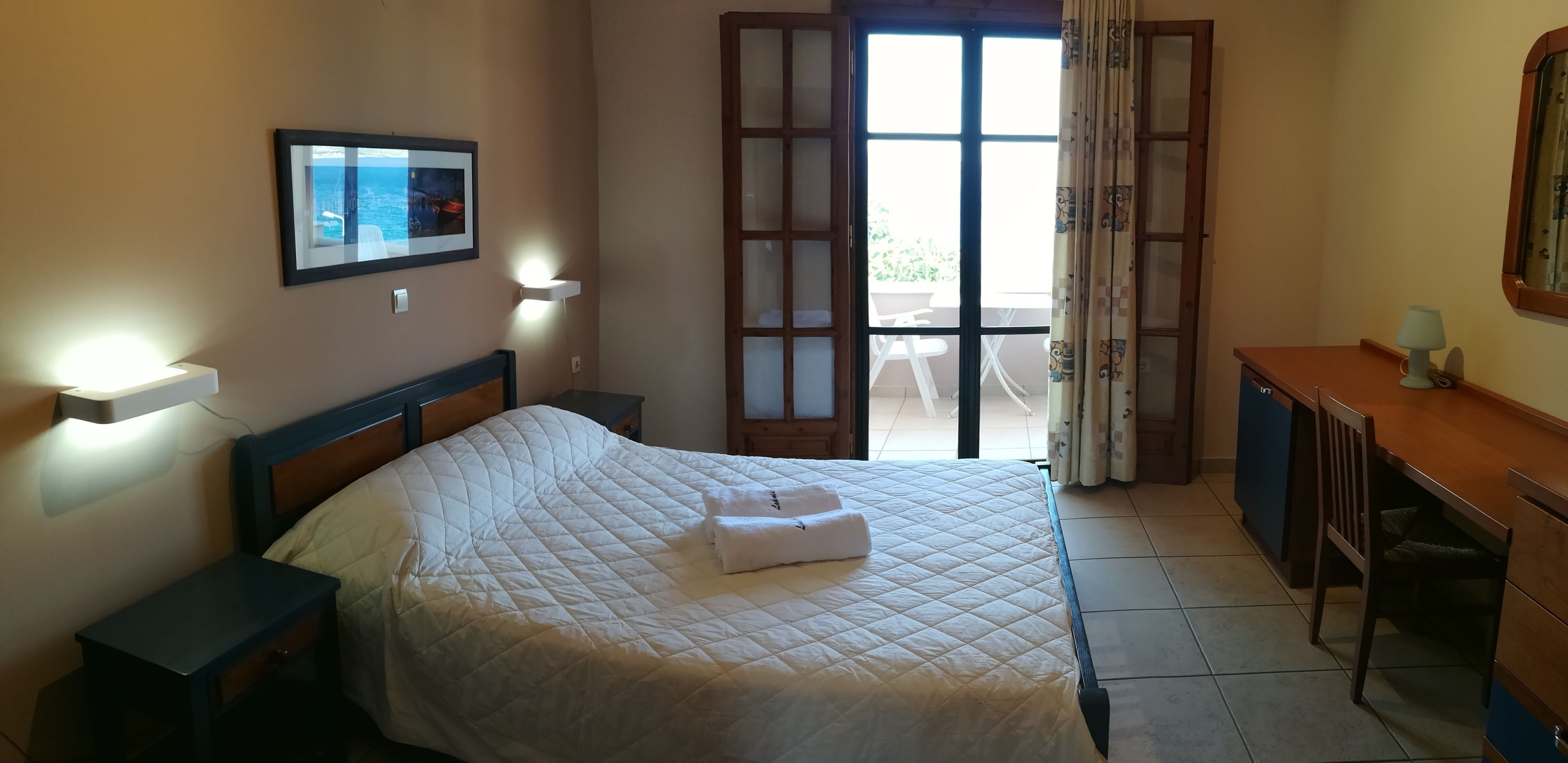 Quarto de hotel com cama, mesa de cabeceira, luminária e vista para o exterior através de uma porta de vidro.