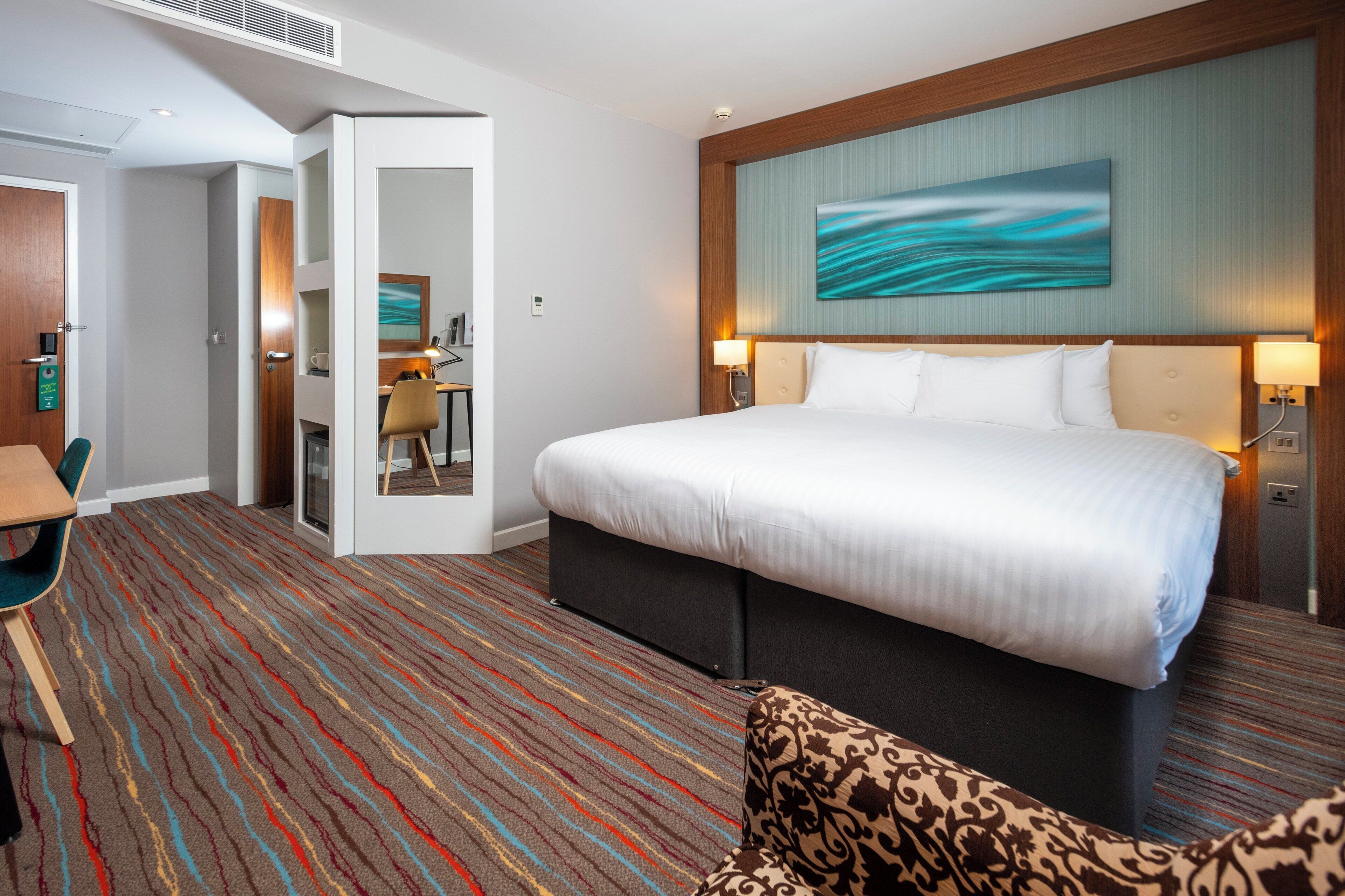 Quarto de hotel com uma cama grande, uma escrivaninha, uma cadeira e um tapete colorido.