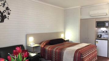 Quarto de hotel com cama, escrivaninha, cadeira, mesa de cabeceira, ar condicionado fixado na parede e cozinha compacta com micro-ondas e geladeira.