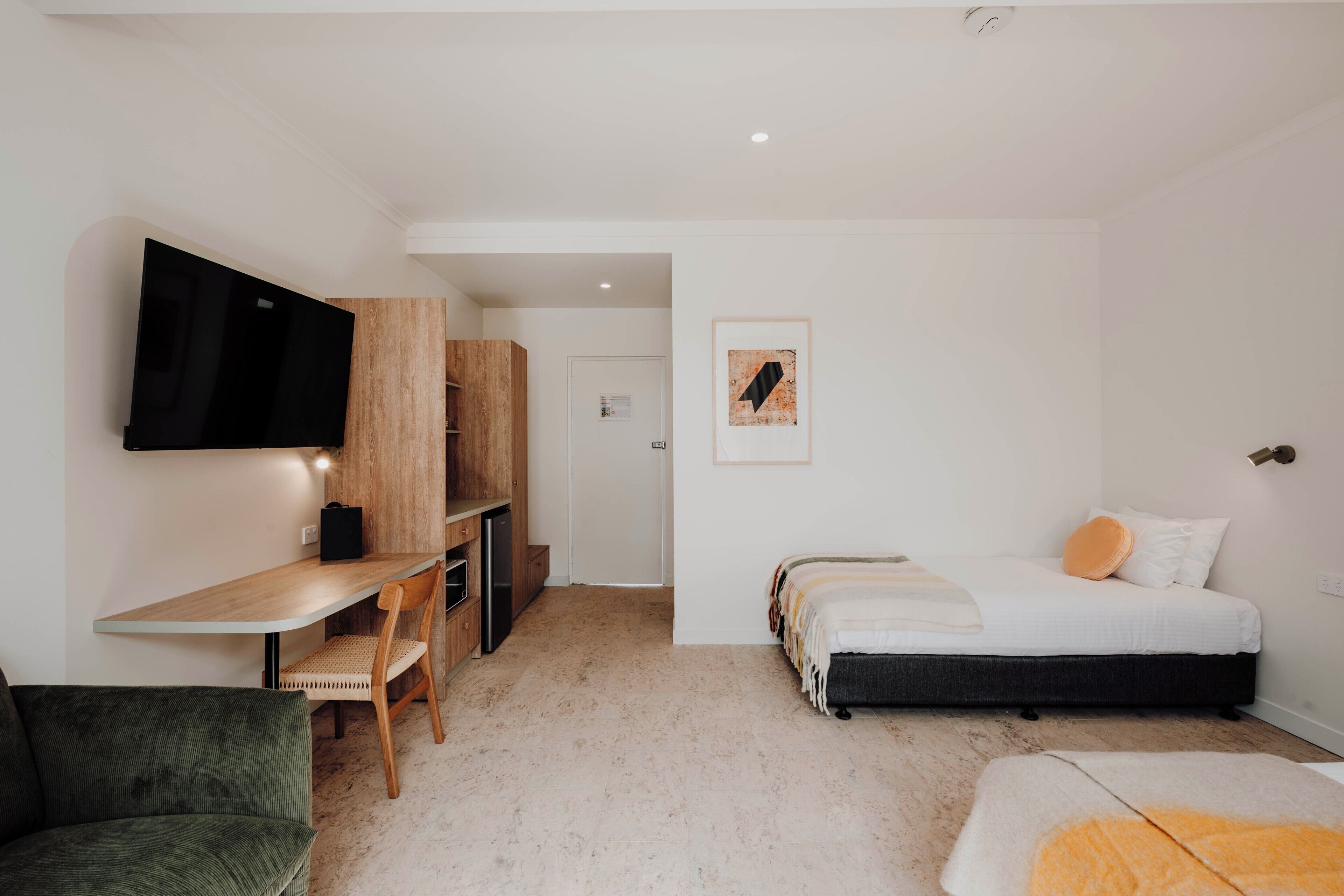 Quarto moderno e compacto, com cama, escrivaninha, cadeira e televisão.