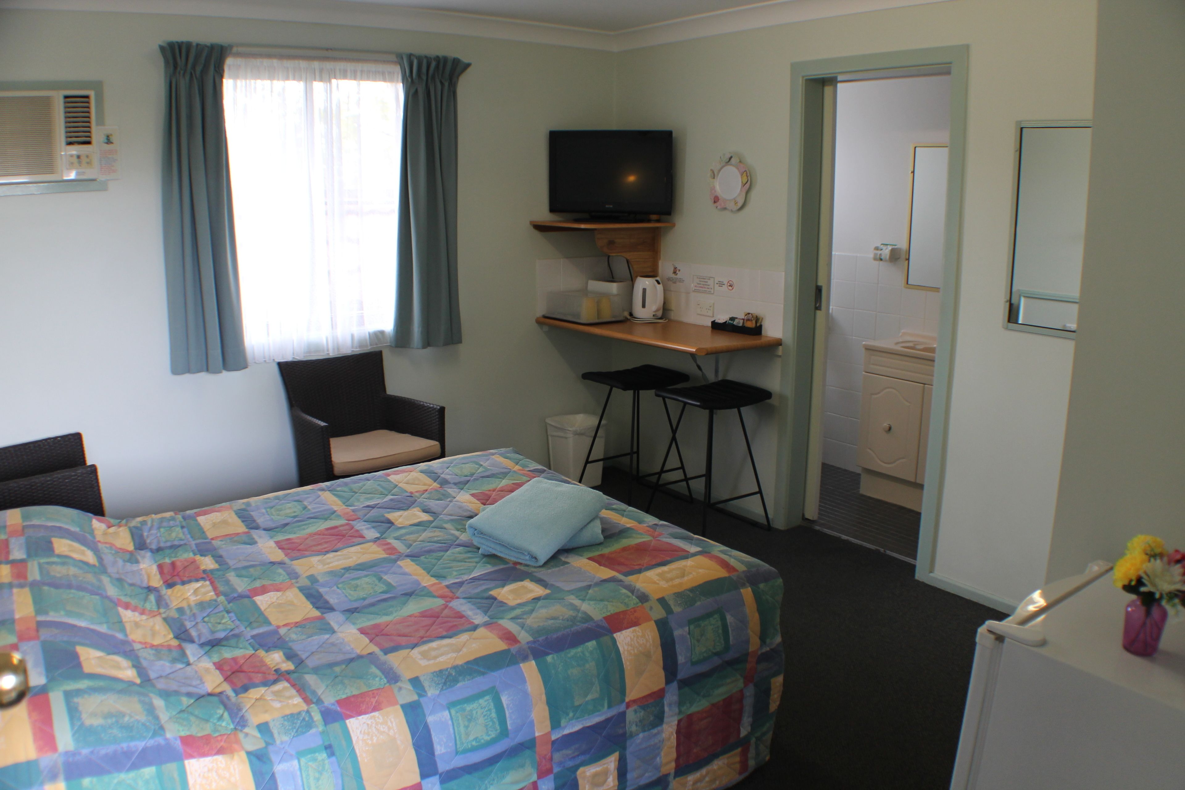 Quarto de hotel com cama, escrivaninha com televisão, uma cadeira e banheiro visível por uma porta aberta.