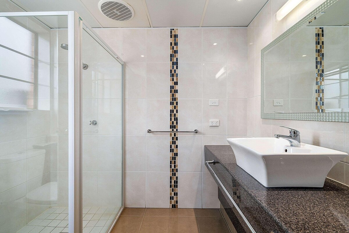 Uma casa de banho moderna com cabine de duche em vidro, lavatório branco e parede revestida com um detalhe decorativo.