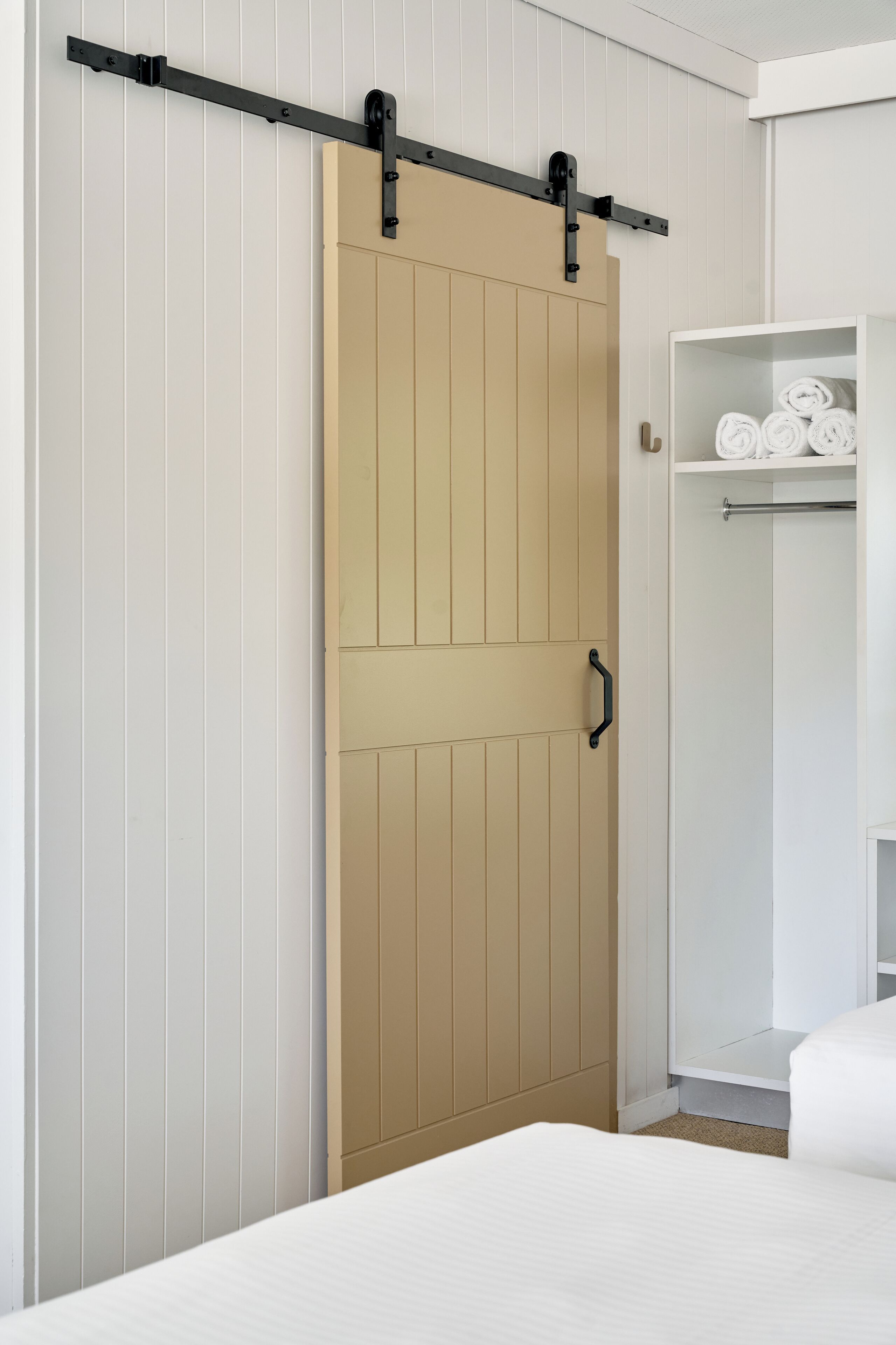 Quarto com porta deslizante, cama branca e guarda-roupa branco com prateleiras.
