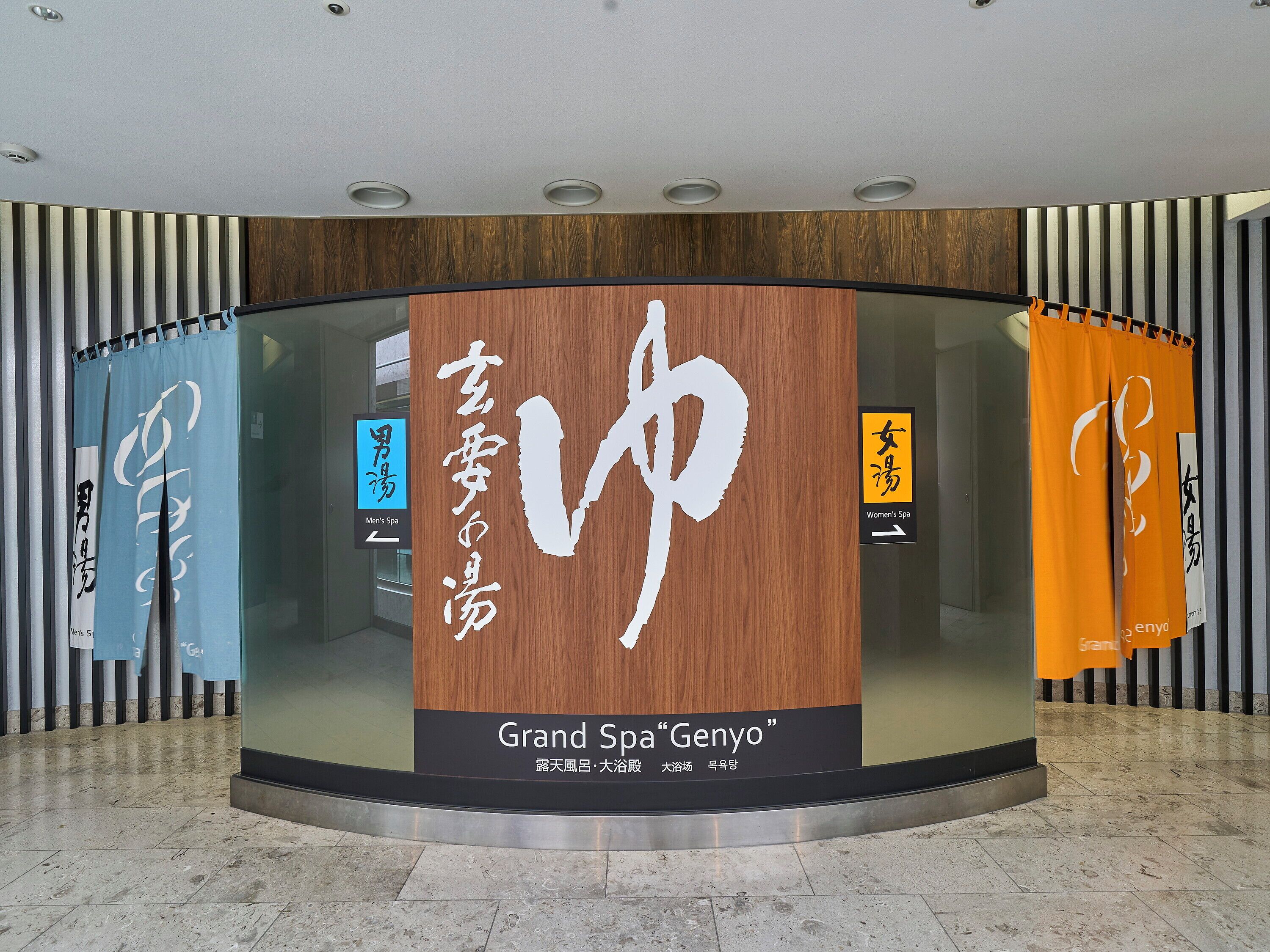 Área de recepção do Grand Spa 'Genyo' com painel de madeira e portas de vidro, placas indicando Spas masculino e feminino, e piso revestido.