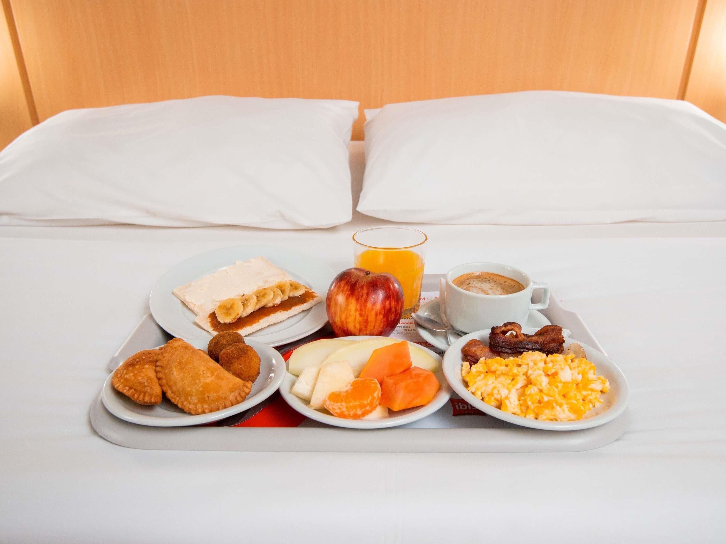 Bandeja de café da manhã com panquecas, fatias de maçã, gomos de laranja, ovos mexidos, croissants e uma xícara de café sobre uma cama de hotel.