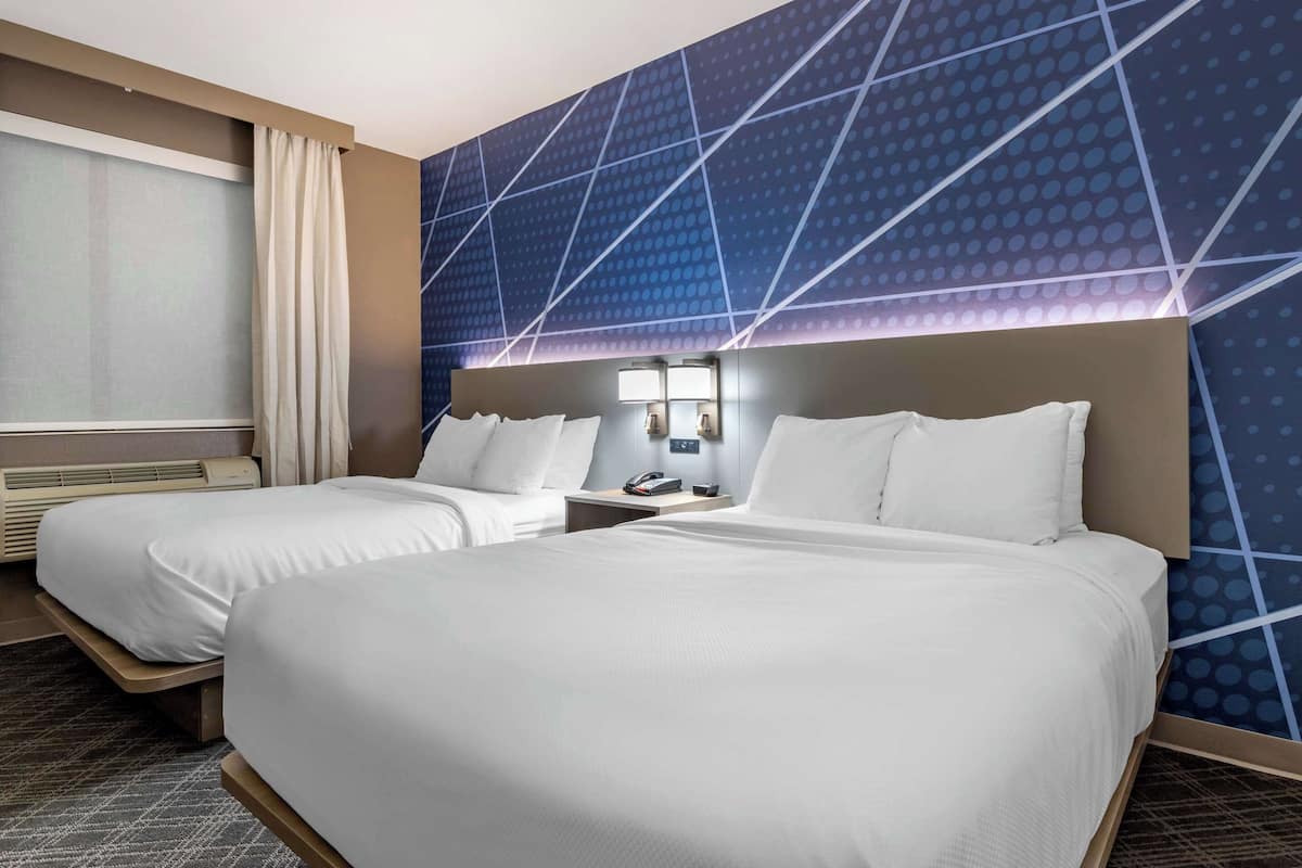 Quarto de hotel com duas camas, uma janela grande com cortinas, parede com padrão geométrico e mesa de cabeceira com telefone e luminária.