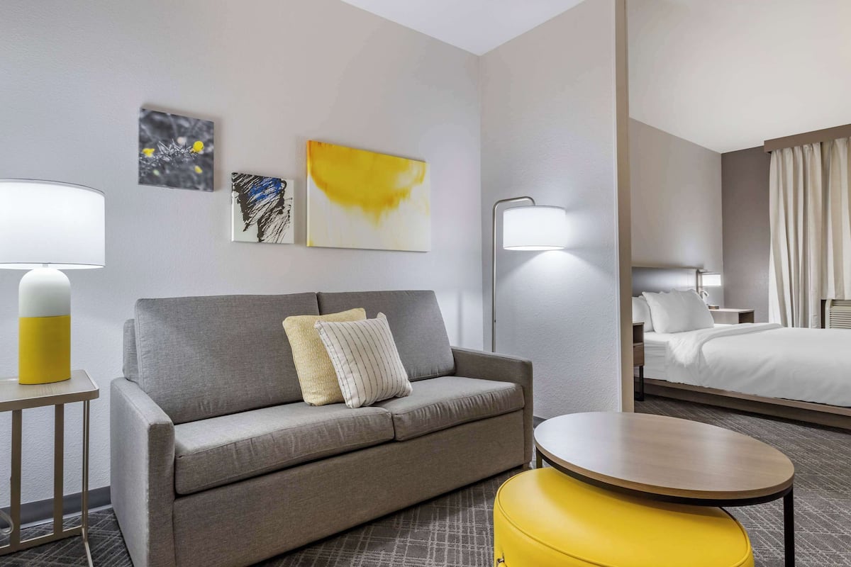 Quarto de hotel moderno com sofá cinza, um banquinho amarelo, mesa de cabeceira, abajur e uma cama com cortinas.