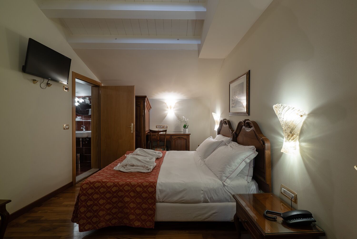 Quarto de hotel com cama, mesa de cabeceira, abajur e televisão na parede.