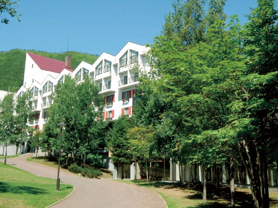 Um hotel moderno de vários andares, com um telhado vermelho, rodeado por vegetação exuberante e uma entrada com caminho sinuoso.