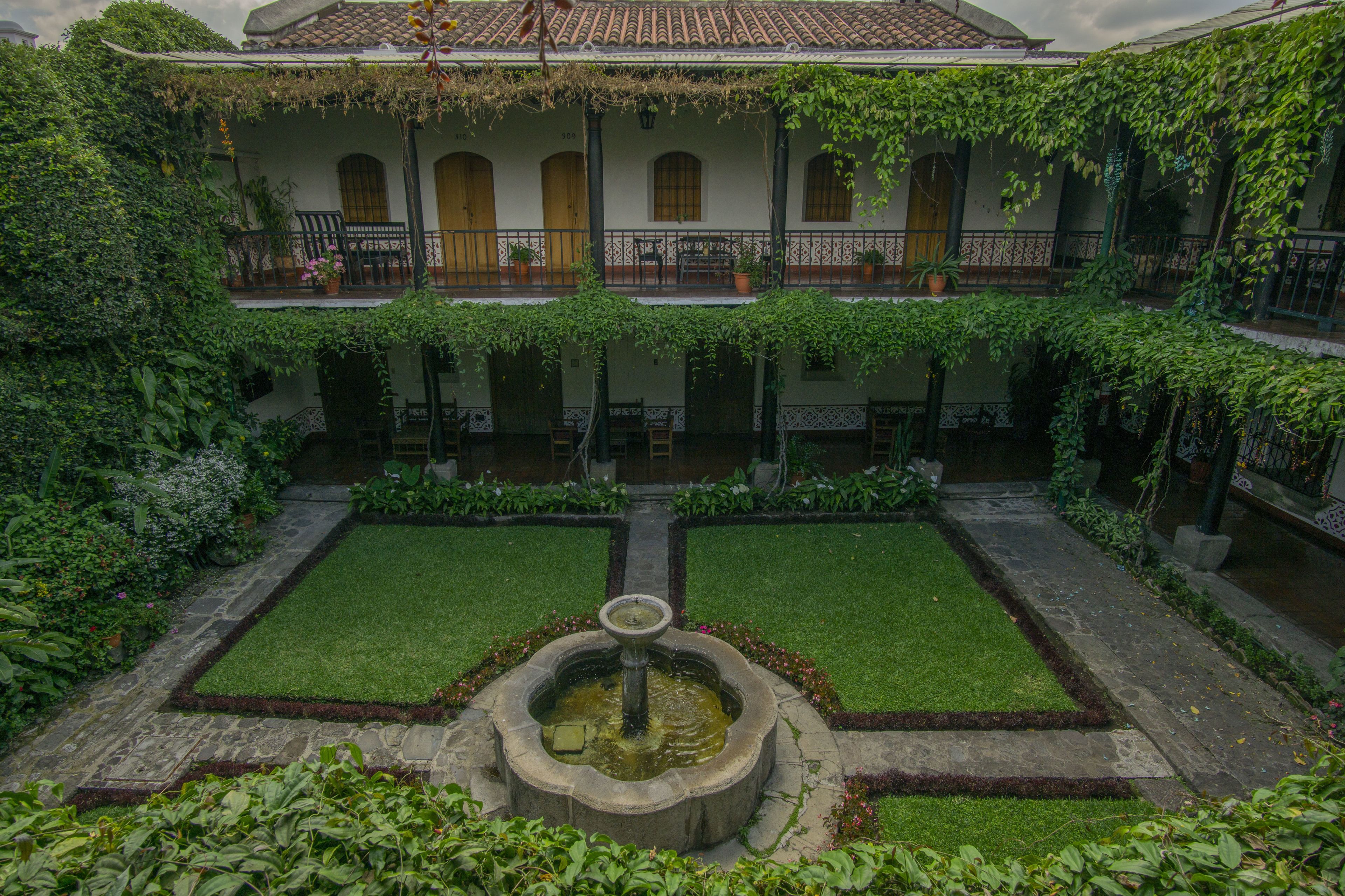 Um pátio com uma fonte central, rodeado por vegetação exuberante e um edifício com várias varandas.