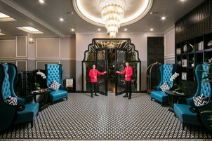 Um hall com piso estampado em preto e branco, poltronas estofadas em azul, um lustre e entrada por porta de vidro.