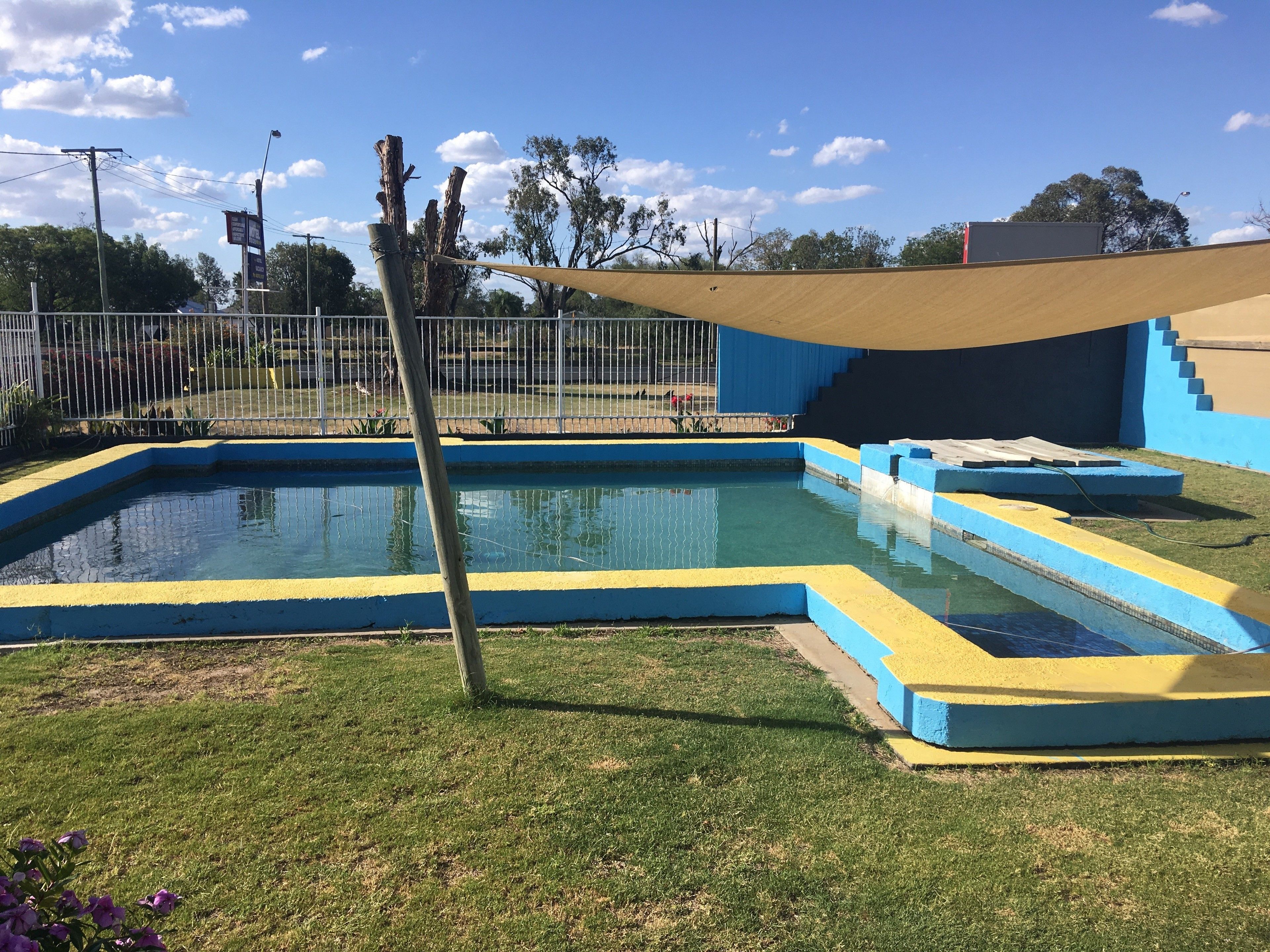 Piscina com bordas azuis e amarelas, cercada por área gramada e árvores.
