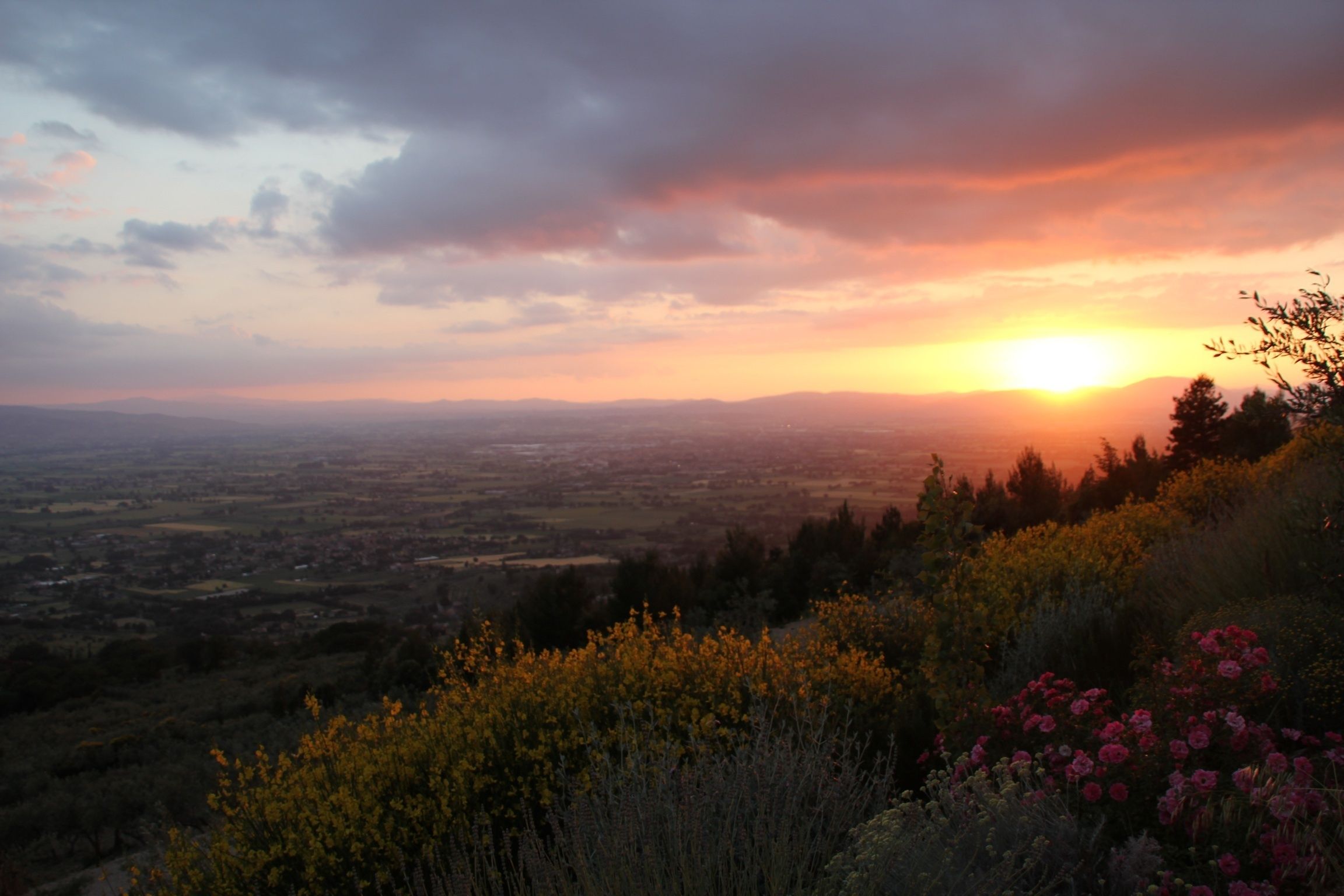 Vista panorâmica de uma paisagem com o pôr do sol sobre um vale, mostrando uma mistura de vegetação natural e uma cidade ao longe.