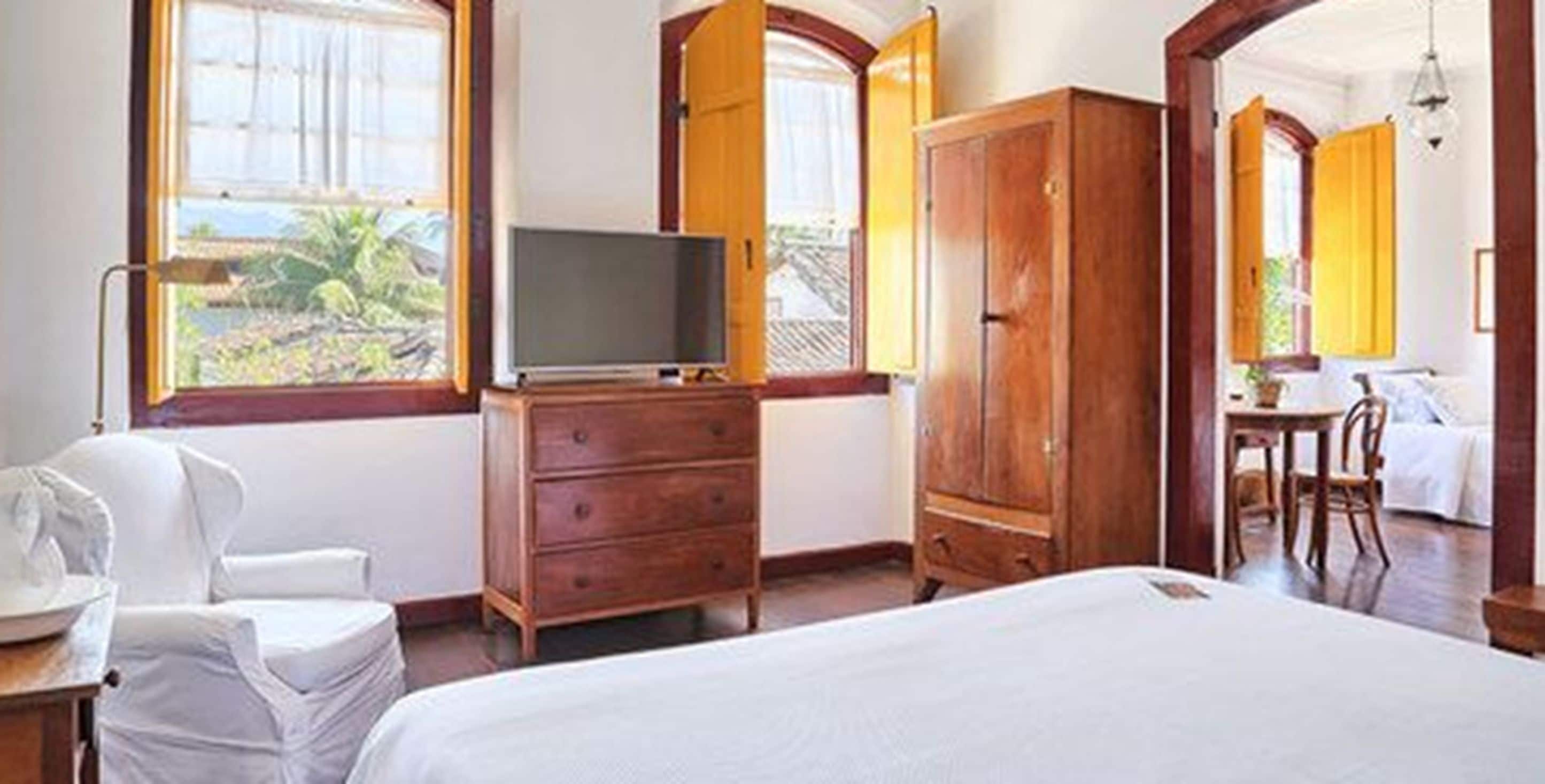 Quarto de hotel com uma cama, cômoda de madeira, televisão e janela com vista para áreas verdes.
